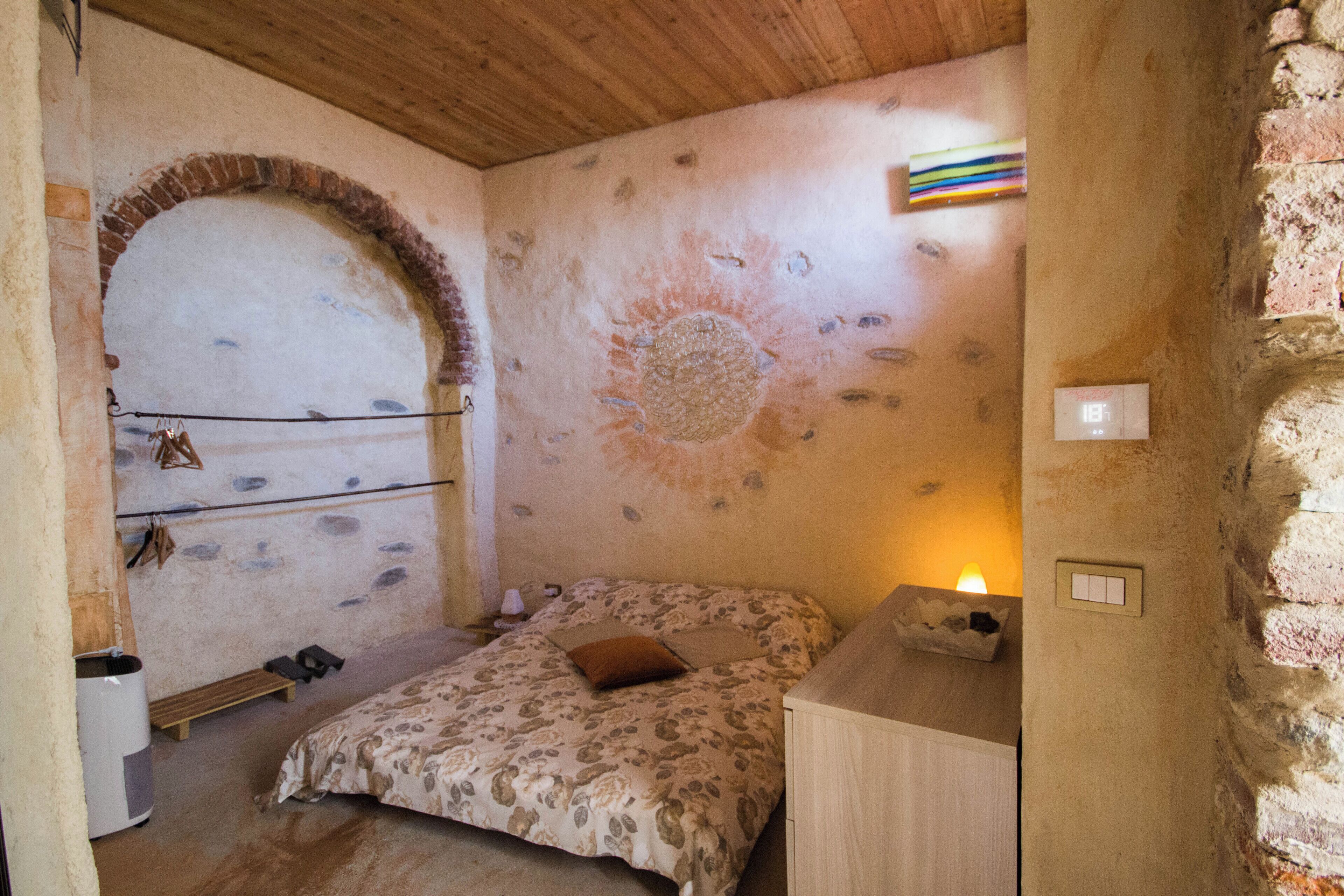Quarto com cama, um banquinho de madeira, uma mesa pequena e uma parede com arco de tijolos.