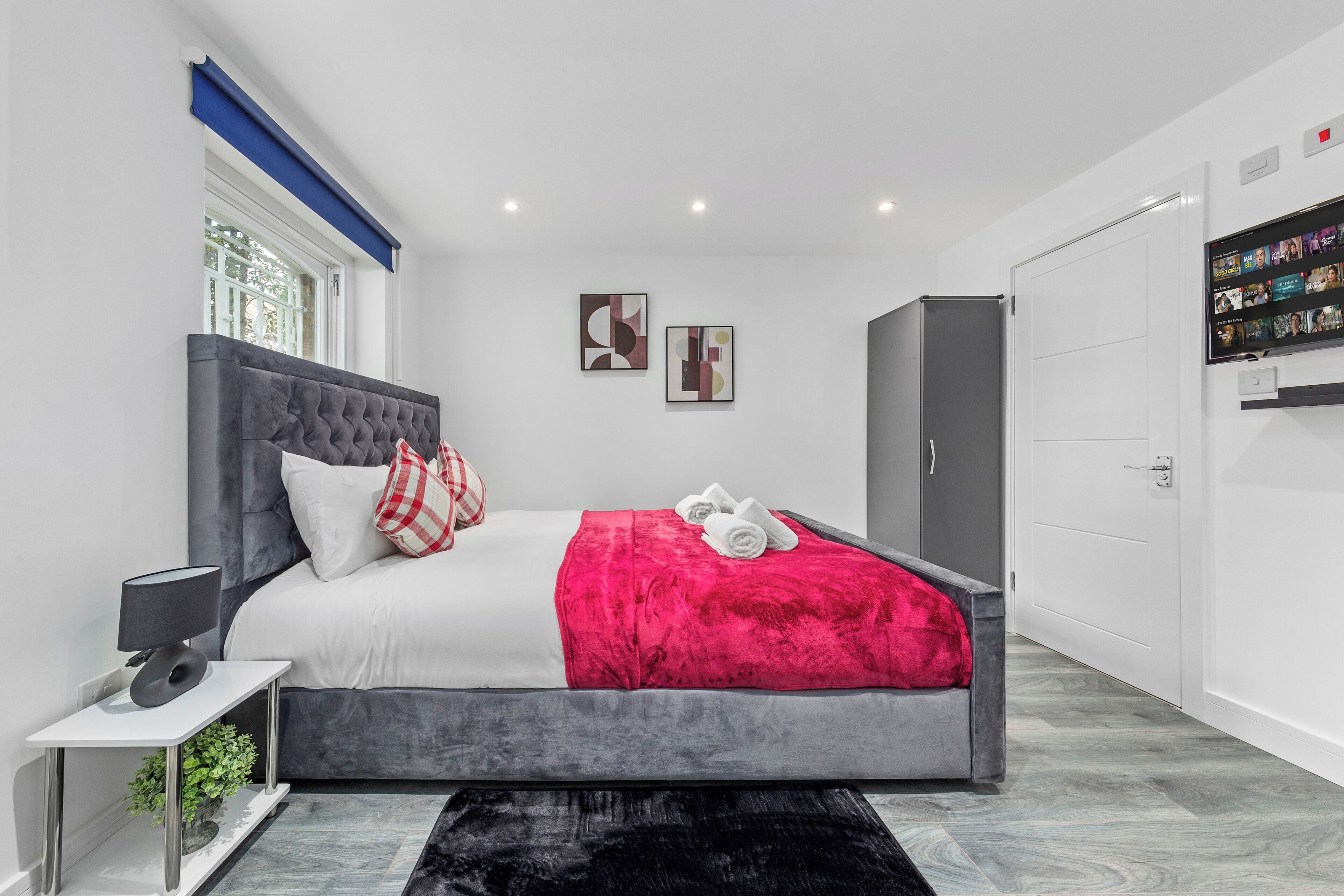 Quarto moderno com cama estofada cinza, roupa de cama branca e manta vermelha. Há uma pequena mesa de cabeceira com uma luminária e uma planta. O quarto tem paredes brancas, uma janela com persianas azuis e um guarda-roupa cinza.
