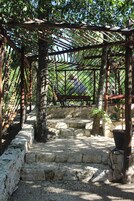 Uma trilha de pedra leva a uma pérgola de madeira com um muro de pedra ao fundo.