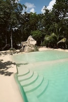 Piscina transparente com formação rochosa e palmeiras ao fundo.