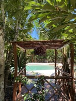 Uma pérgola de madeira com decoração pendente, piscina e vegetação tropical ao redor.