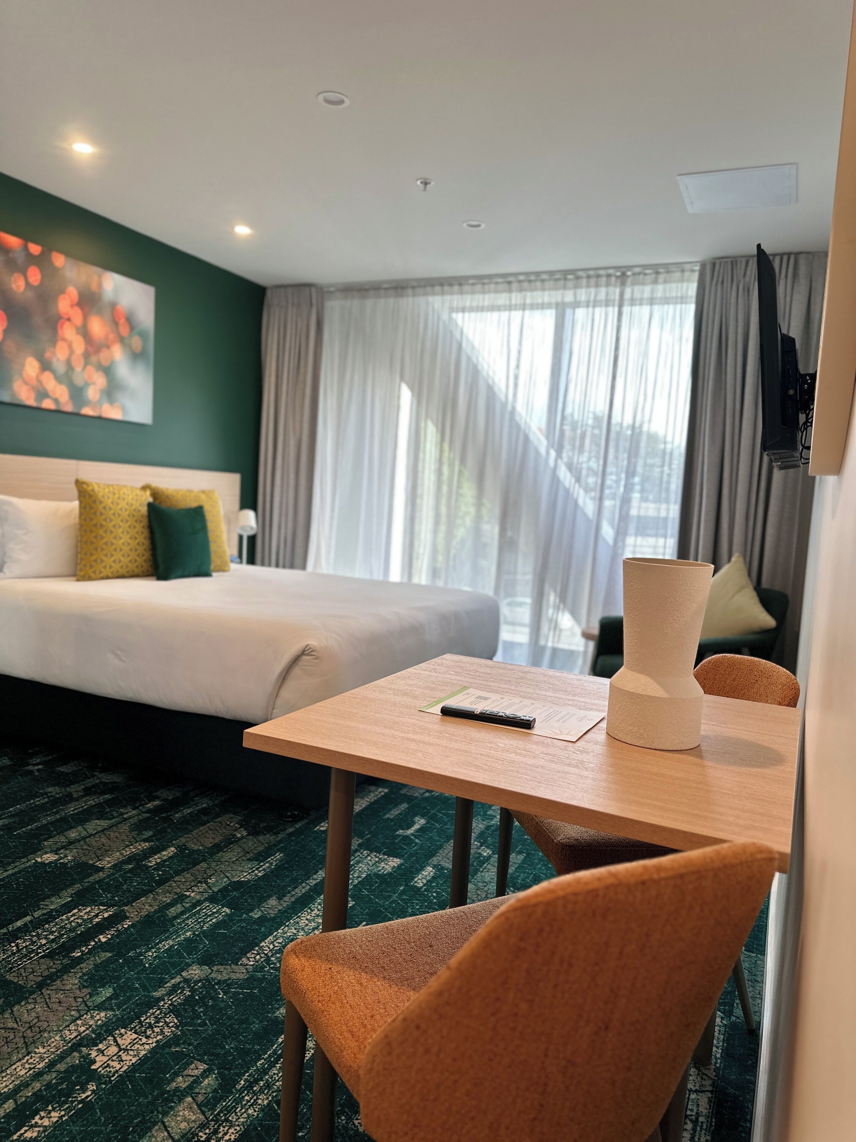 Quarto de hotel com uma cama, uma mesa com controle remoto, uma cadeira e uma televisão presa na parede.