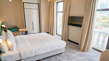 Quarto de hotel com uma cama grande, uma mesinha redonda, um sofá, uma televisão na parede e vista para o exterior pela janela.