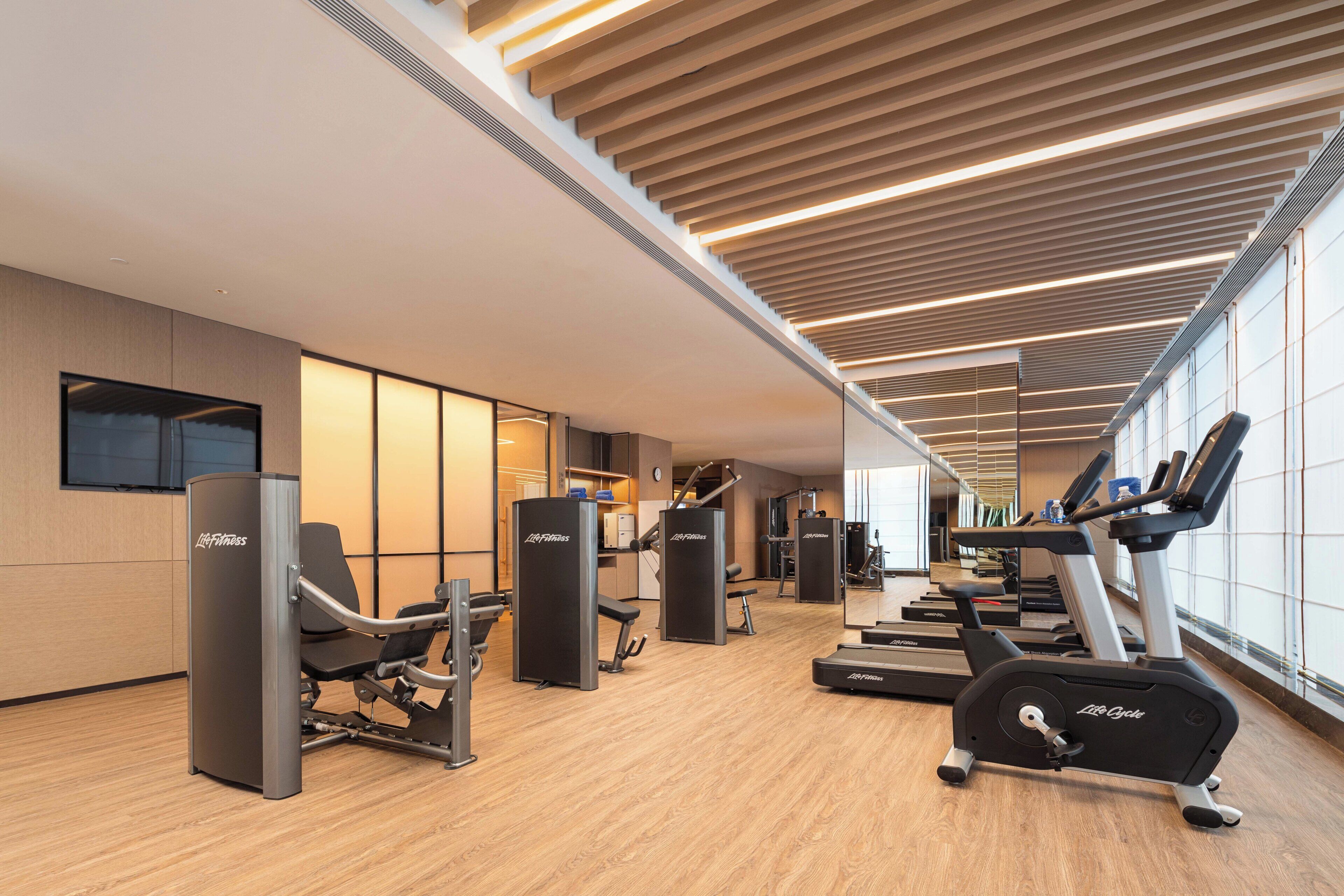 Academia moderna com máquinas de cardio, estações de musculação e piso de madeira.