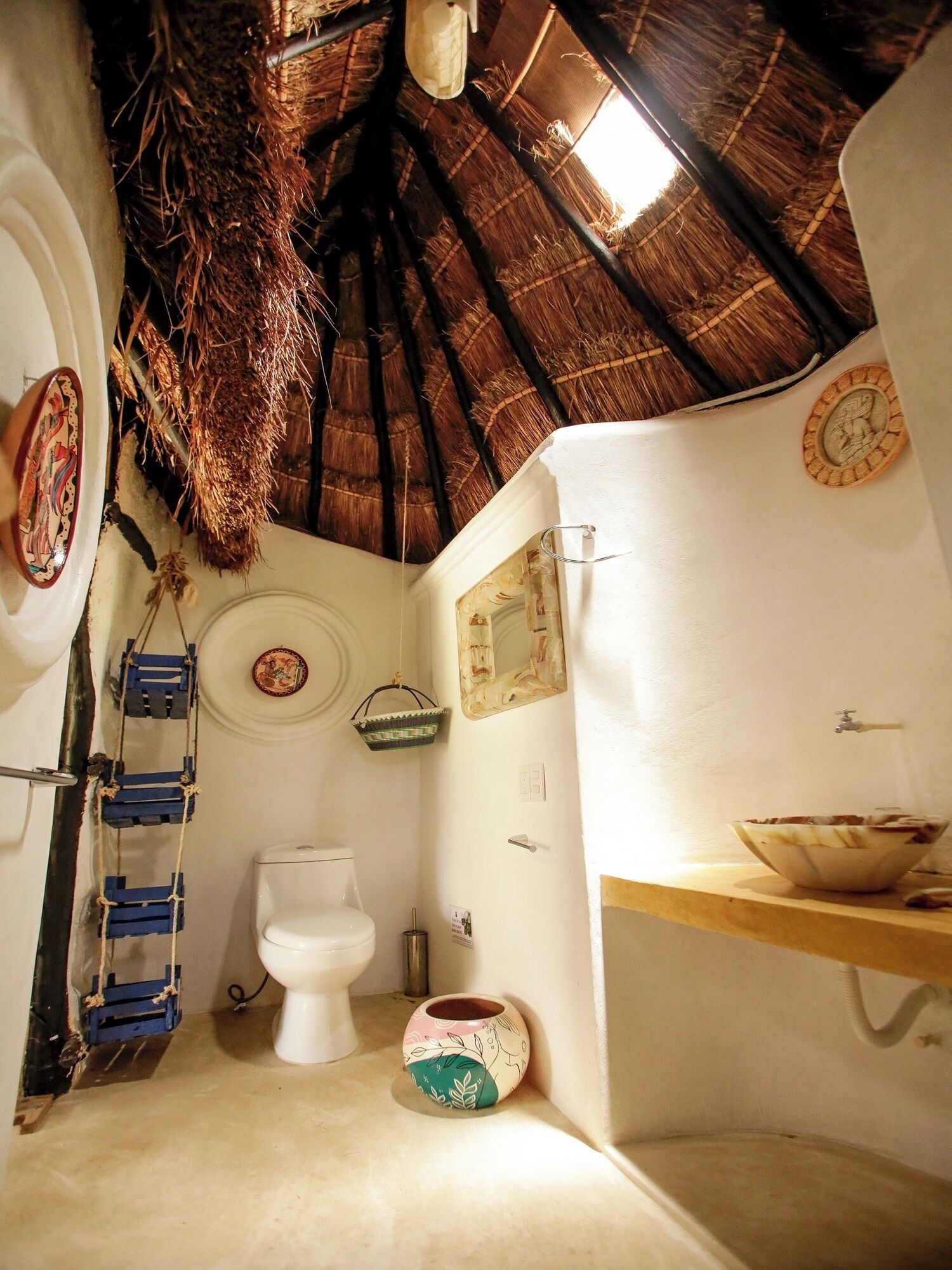 Um banheiro compacto com telhado de palha, um vaso sanitário, uma pia e uma tigela decorativa.