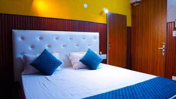 Quarto de hotel com uma cama grande, dois travesseiros azuis, um edredom branco e um guarda-roupa de madeira.