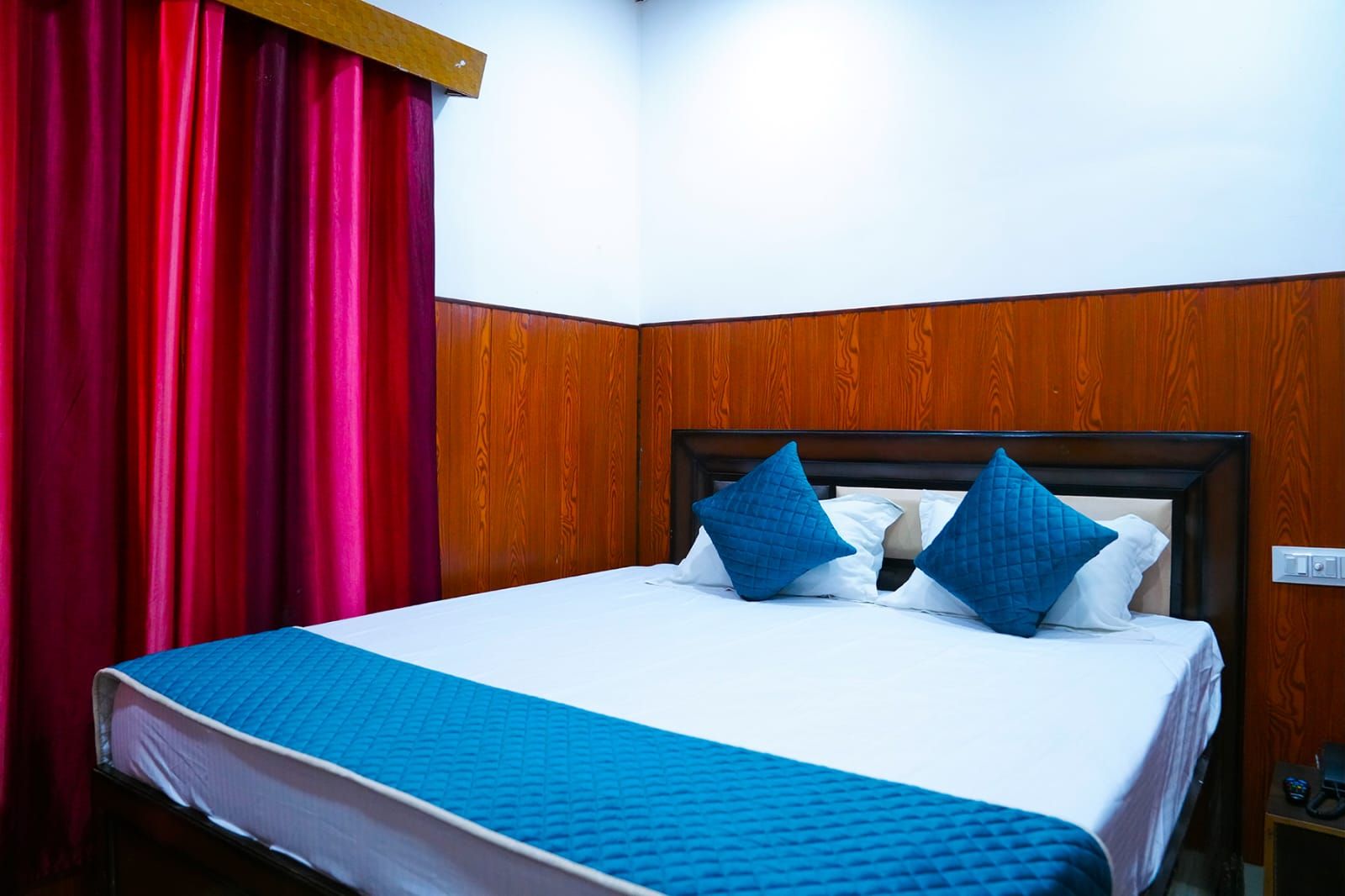 Uma cama bem arrumada com roupa de cama e almofadas azuis, uma cabeceira de madeira e uma cortina vermelha à esquerda.