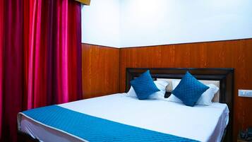 Uma cama bem arrumada com roupa de cama e almofadas azuis, uma cabeceira de madeira e uma cortina vermelha à esquerda.