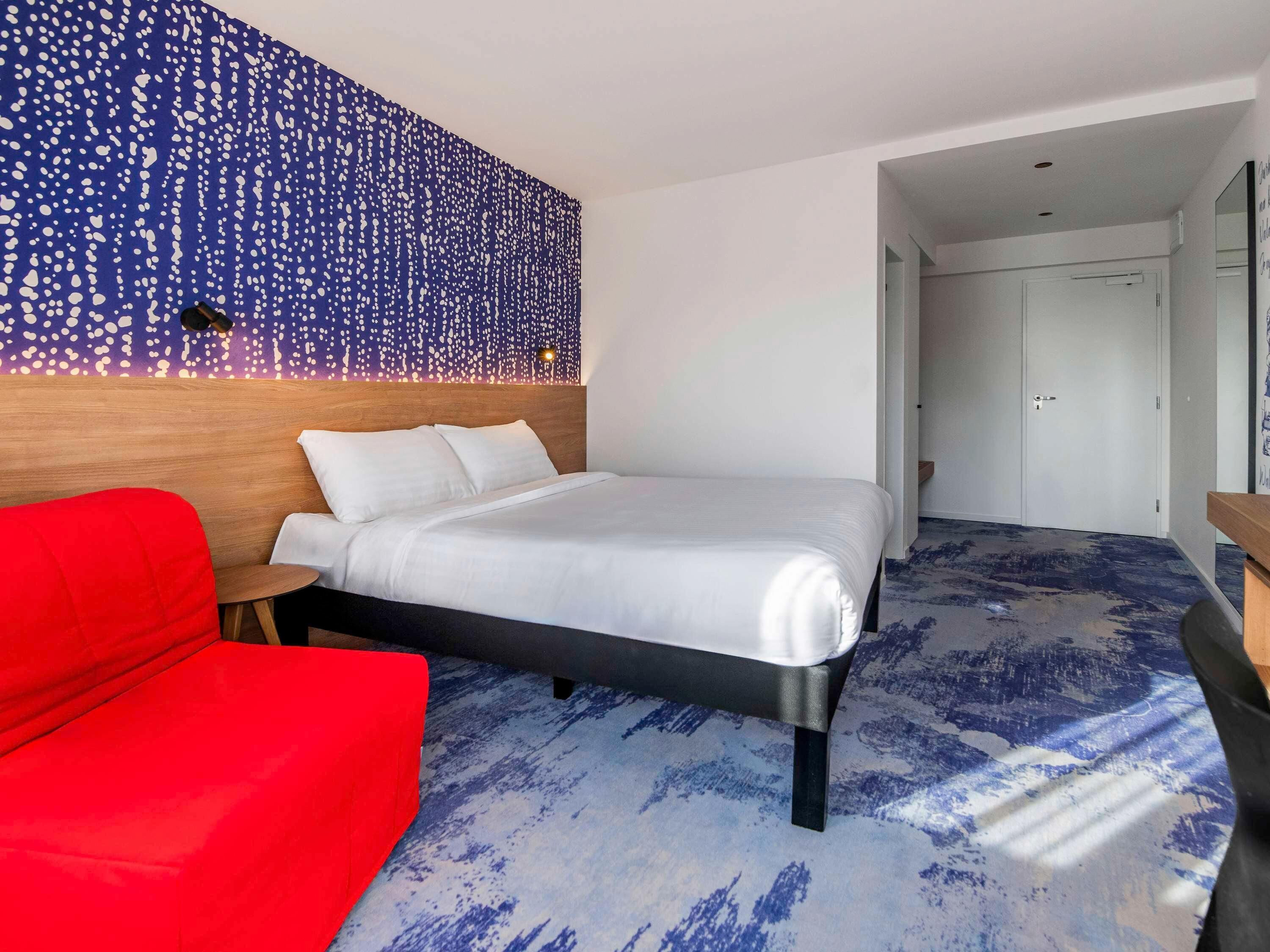 Quarto de hotel com uma cama, uma cadeira vermelha, um painel de cabeceira de madeira e uma parede com padrão azul e branco.