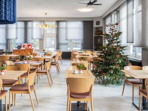 Uma sala de jantar moderna com mesas e cadeiras de madeira, um pinheiro de Natal e vista para o exterior através de grandes janelas.