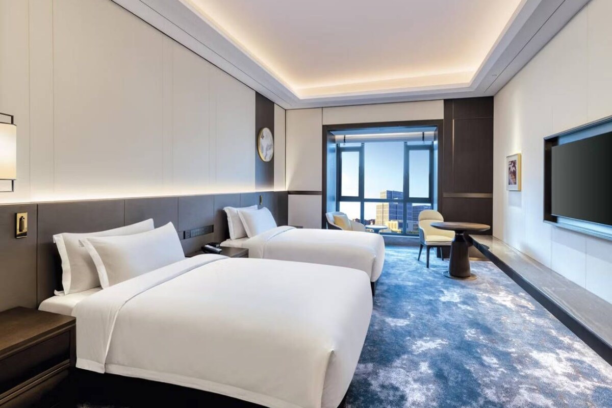 Quarto de hotel com duas camas, uma escrivaninha, uma cadeira e uma janela grande com vista para a cidade.