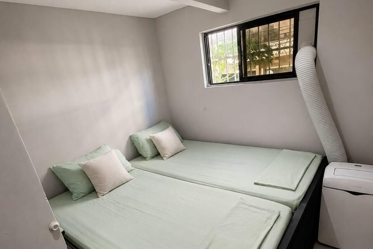 Quarto pequeno e moderno com duas camas, uma janela com persianas, um radiador branco e uma secretária.