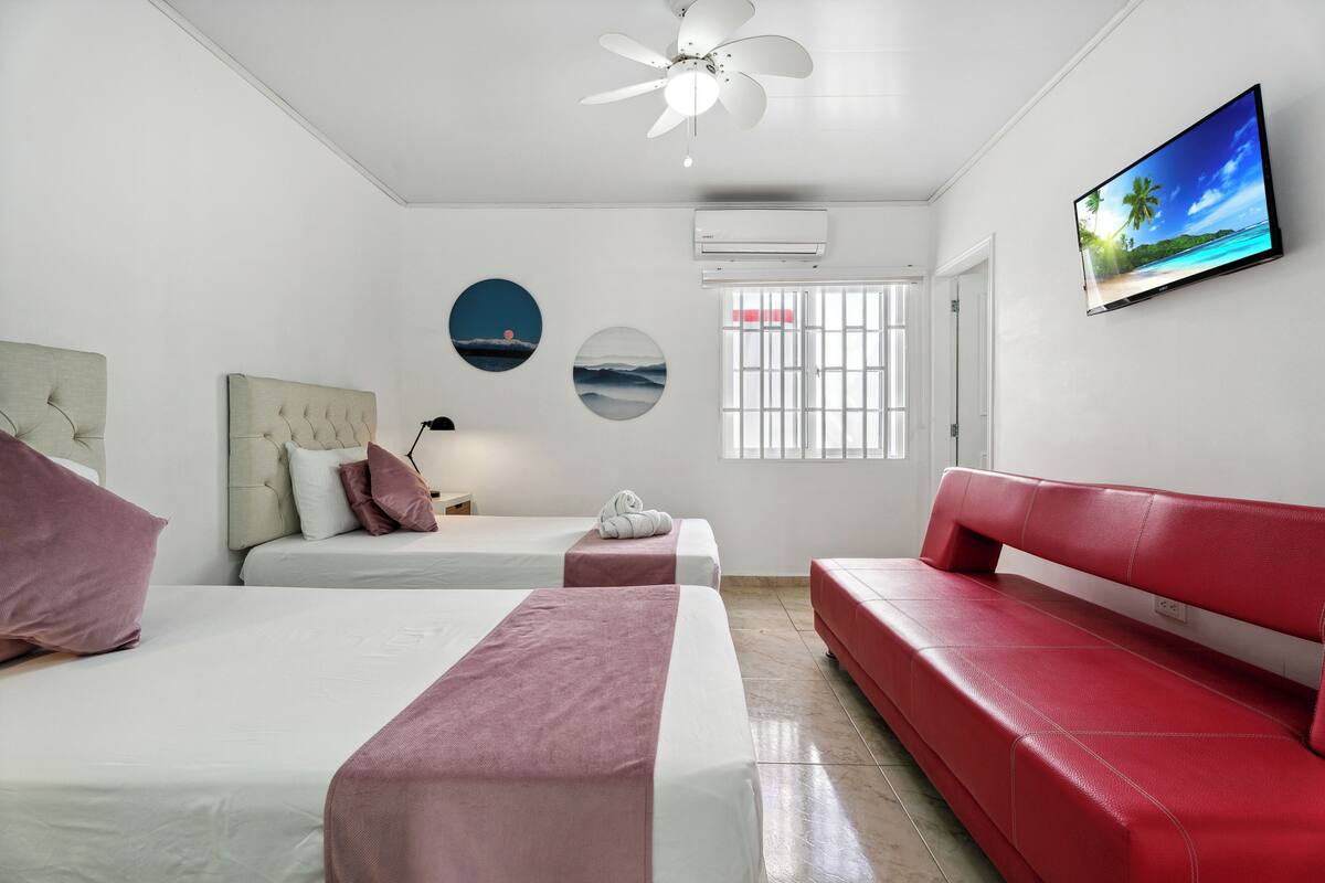 Quarto de hotel com duas camas, um sofá vermelho, ventoinha no teto, televisão na parede e uma janela com grades.
