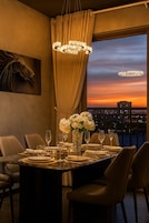 Uma sala de jantar com uma mesa posta para quatro pessoas, um vaso de flores brancas e vista para a cidade ao pôr do sol.