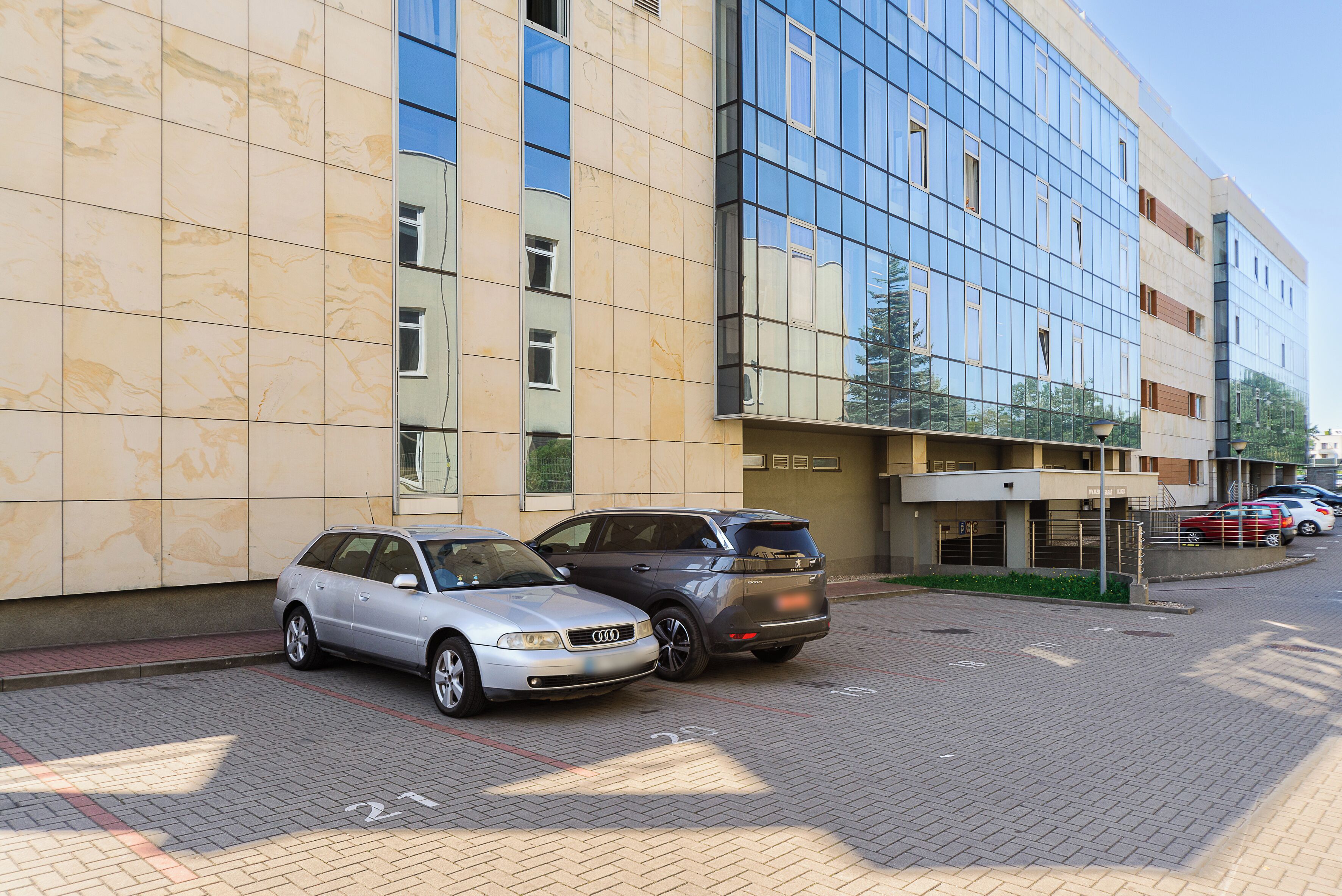 Um edifício moderno com fachada de vidro refletivo e dois carros estacionados na calçada.