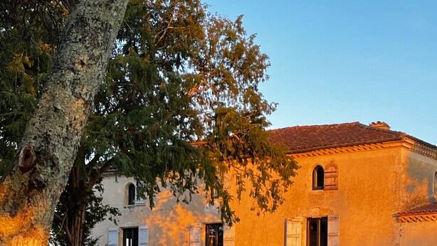 Um edifício rústico com fachada de pedra, várias janelas com persianas e um jardim bem cuidado, com grama e plantas.