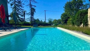 Piscina azul límpida com espreguiçadeiras e guarda-sóis à esquerda, rodeada por vegetação e árvores.