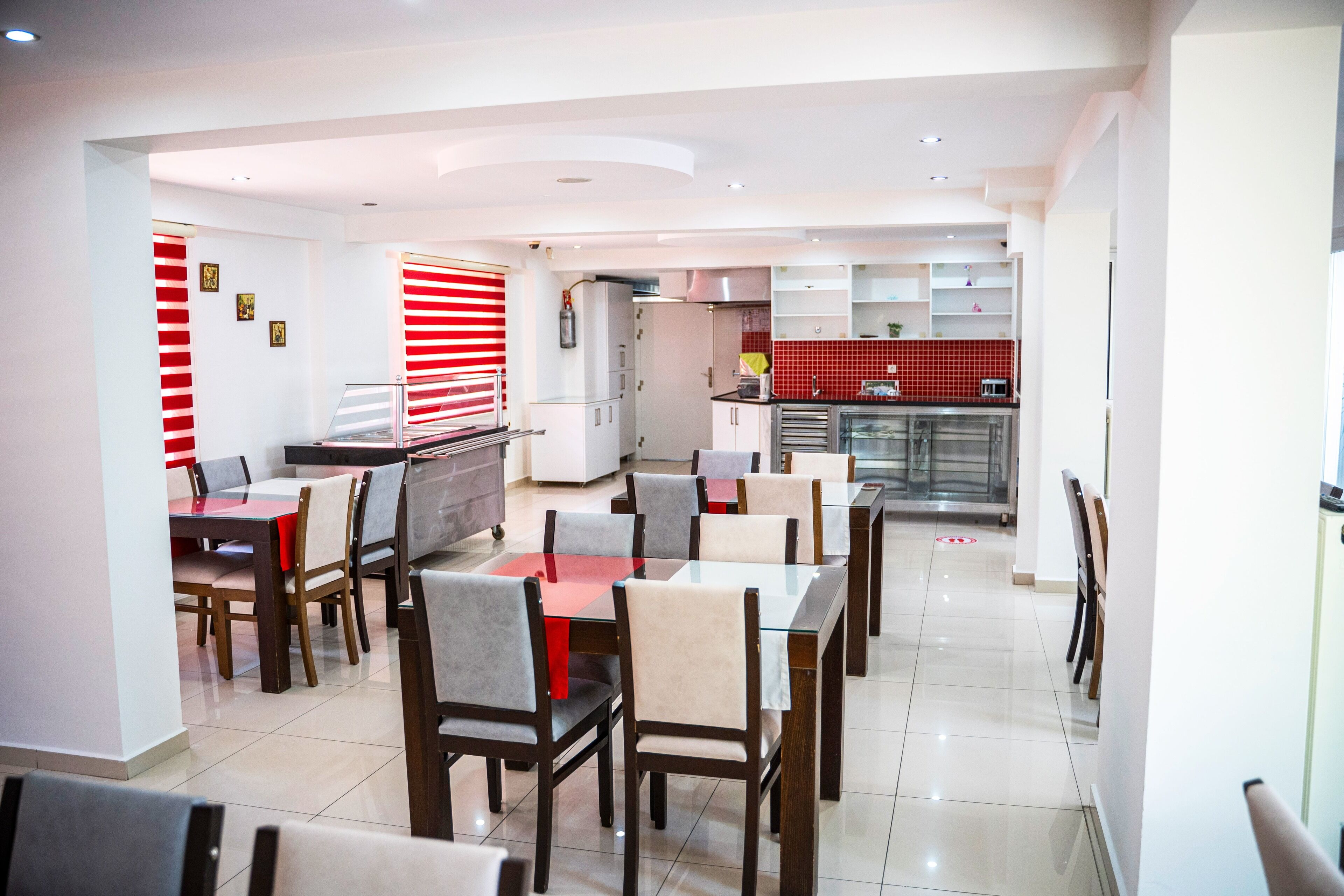 Uma área de refeições moderna com mesas e cadeiras de vidro, uma cozinha com azulejos vermelhos e um interior limpo e claro.