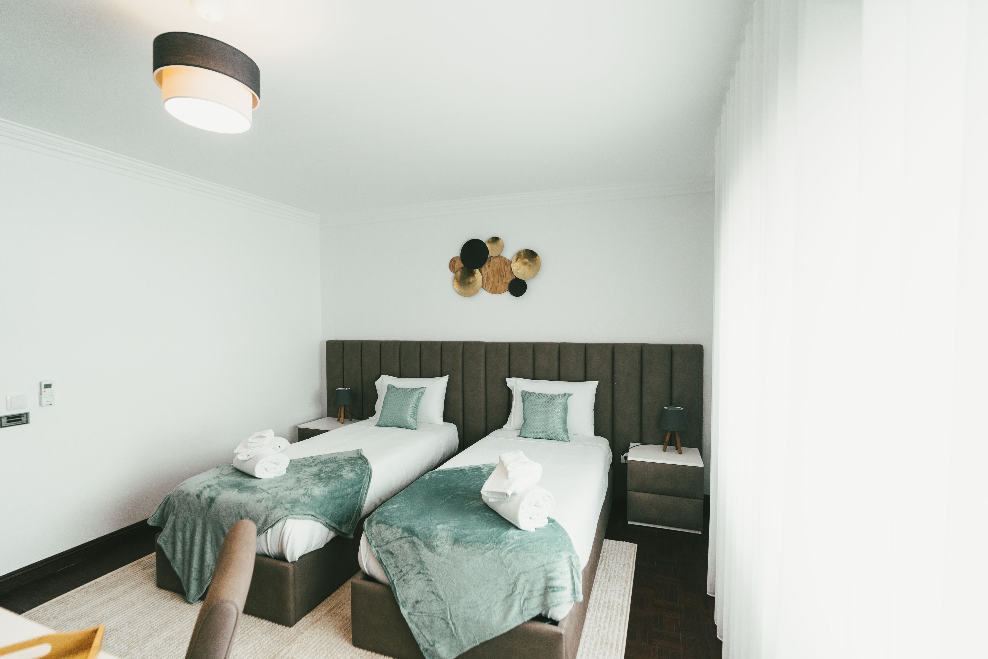 Quarto de hotel moderno com duas camas, um painel de cabeceira escuro e uma peça decorativa sobre as camas.