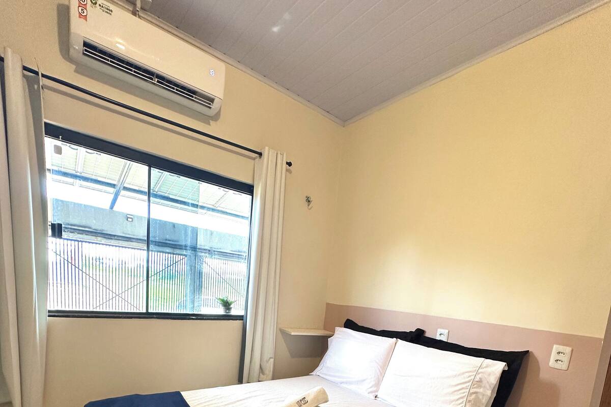 Quarto de hotel com cama, janela com cortinas, aparelho de ar condicionado e vista para o exterior.