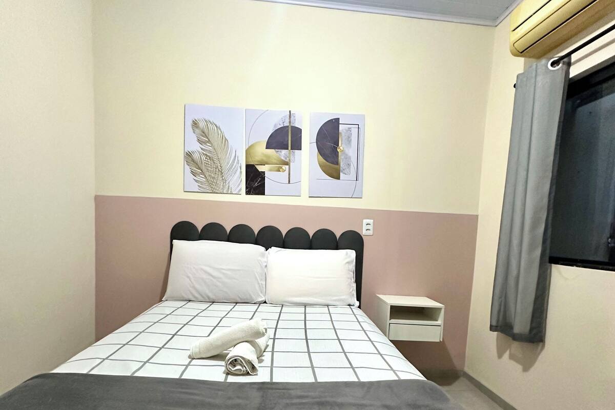 Cama bem arrumada com colcha quadricula preta e branca, manta cinza e duas toalhas enroladas. Três obras de arte estão penduradas na parede acima da cama. Um criado-mudo com abajur fica à direita da cama.