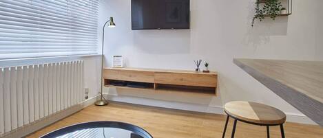 Uma sala de estar moderna com uma mesa redonda de vidro, dois bancos de madeira, uma TV na parede, um aparador de madeira e uma planta em uma prateleira.