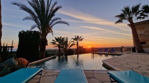 Área de piscina com espreguiçadeiras, palmeiras e vista para o pôr do sol.