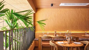 Restaurante com soalho de madeira, teto alto e corrimão decorado com plantas. Mesas preparadas com copos de vinho e um vaso.