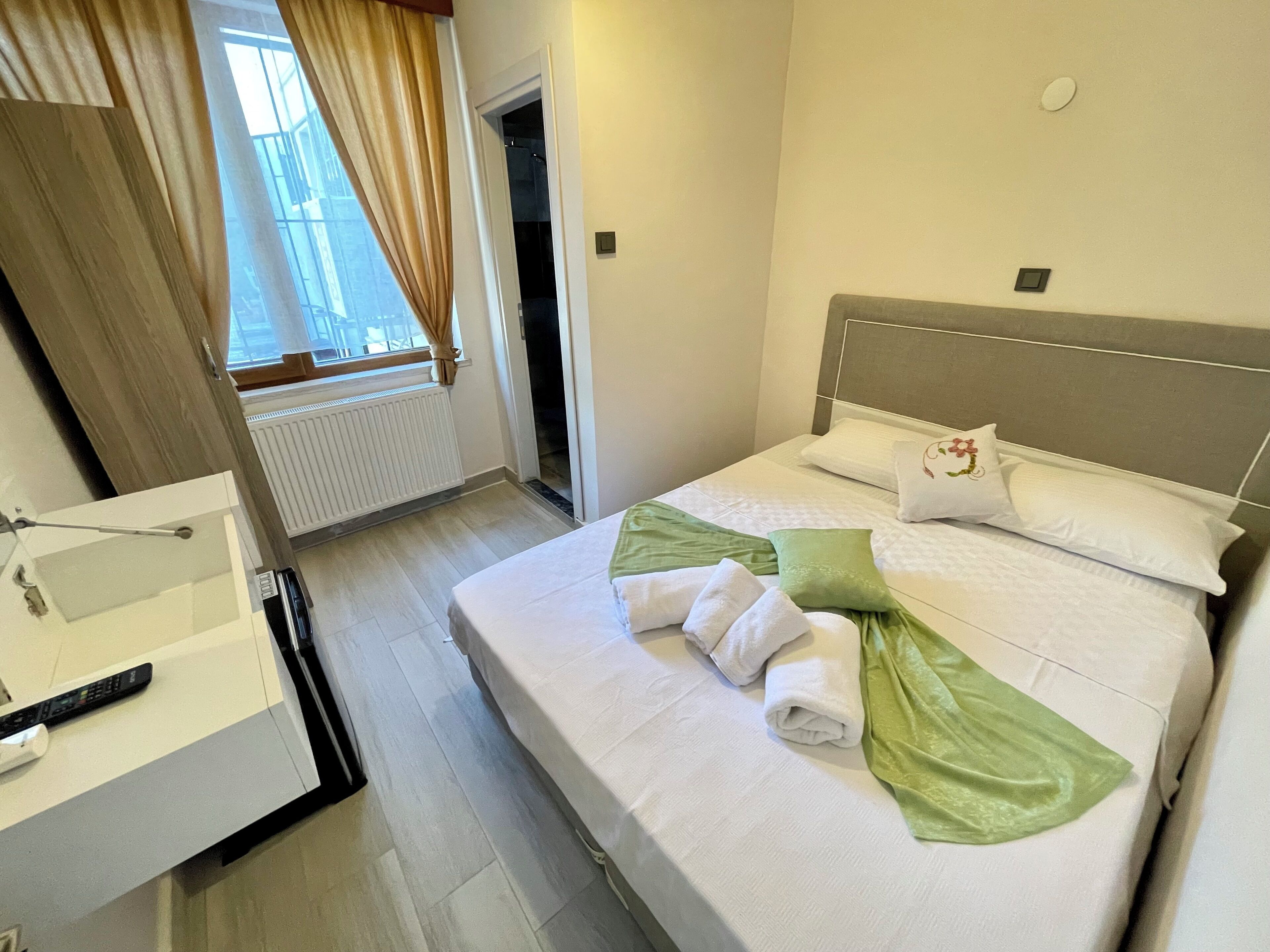 Quarto de hotel com cama, mesa de cabeceira, janela com cortinas e radiador.