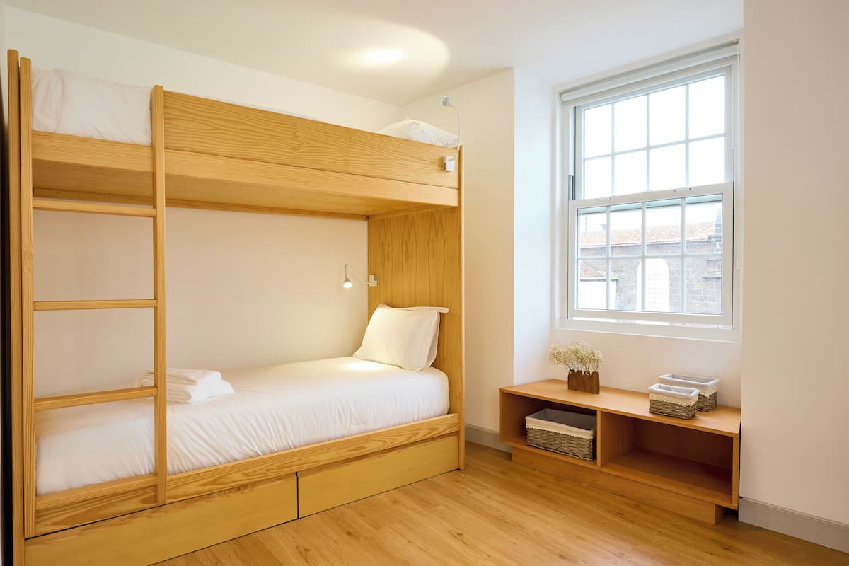 Uma cama beliche com estrutura de madeira, uma mesa de cabeceira com cestos de armazenamento e uma janela com vista para edifícios.