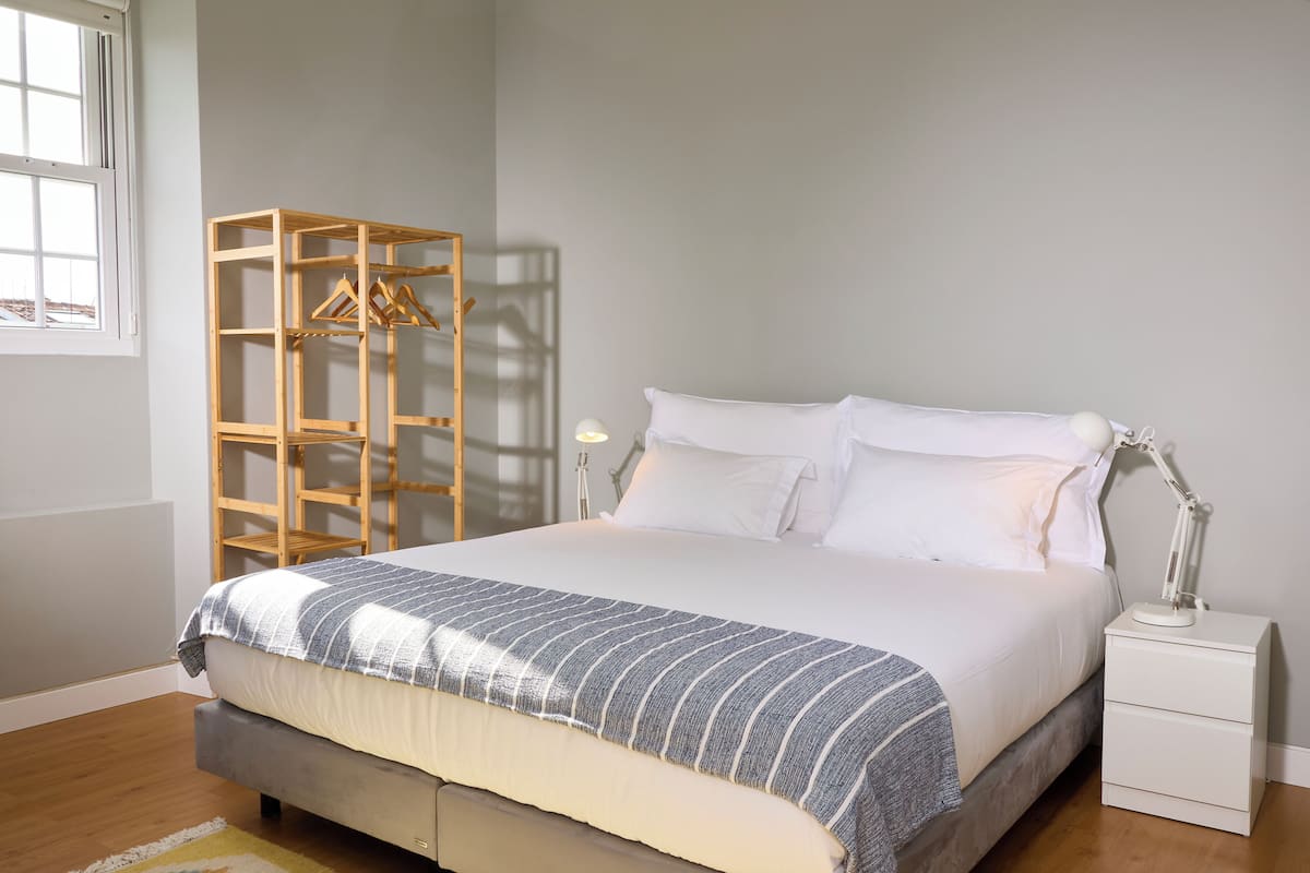 Quarto com cama, mesa de cabeceira com abajur, guarda-roupa de madeira e janela.