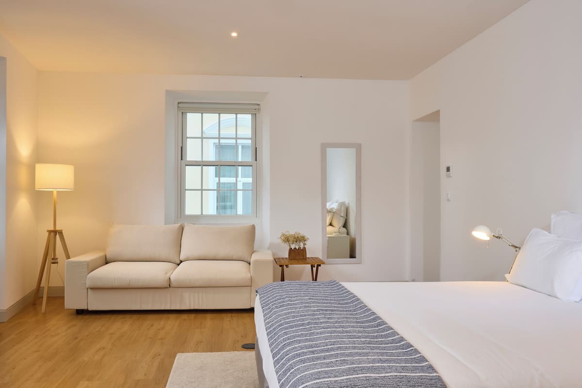 Quarto de hotel moderno com uma cama grande, um sofá, uma mesa pequena com abajur, uma janela e um espelho.