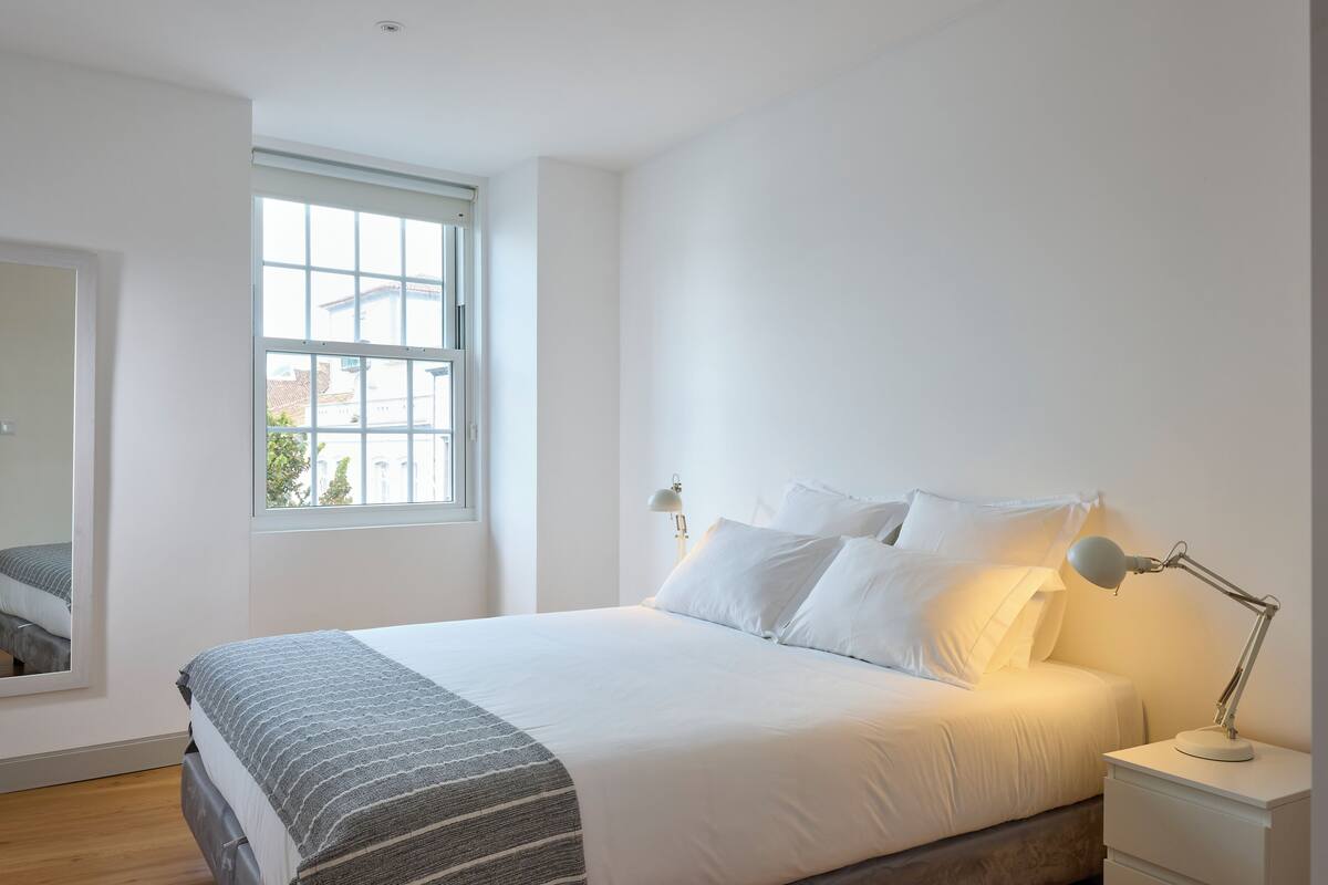Quarto com cama grande, roupa de cama branca, mesa de cabeceira com abajur e janela com vista para edifícios.