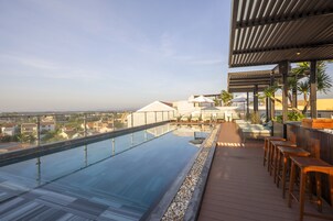 Piscina infinita no terraço com vista ampla da paisagem urbana, cadeiras de espreguiçadeira e área de estar coberta.