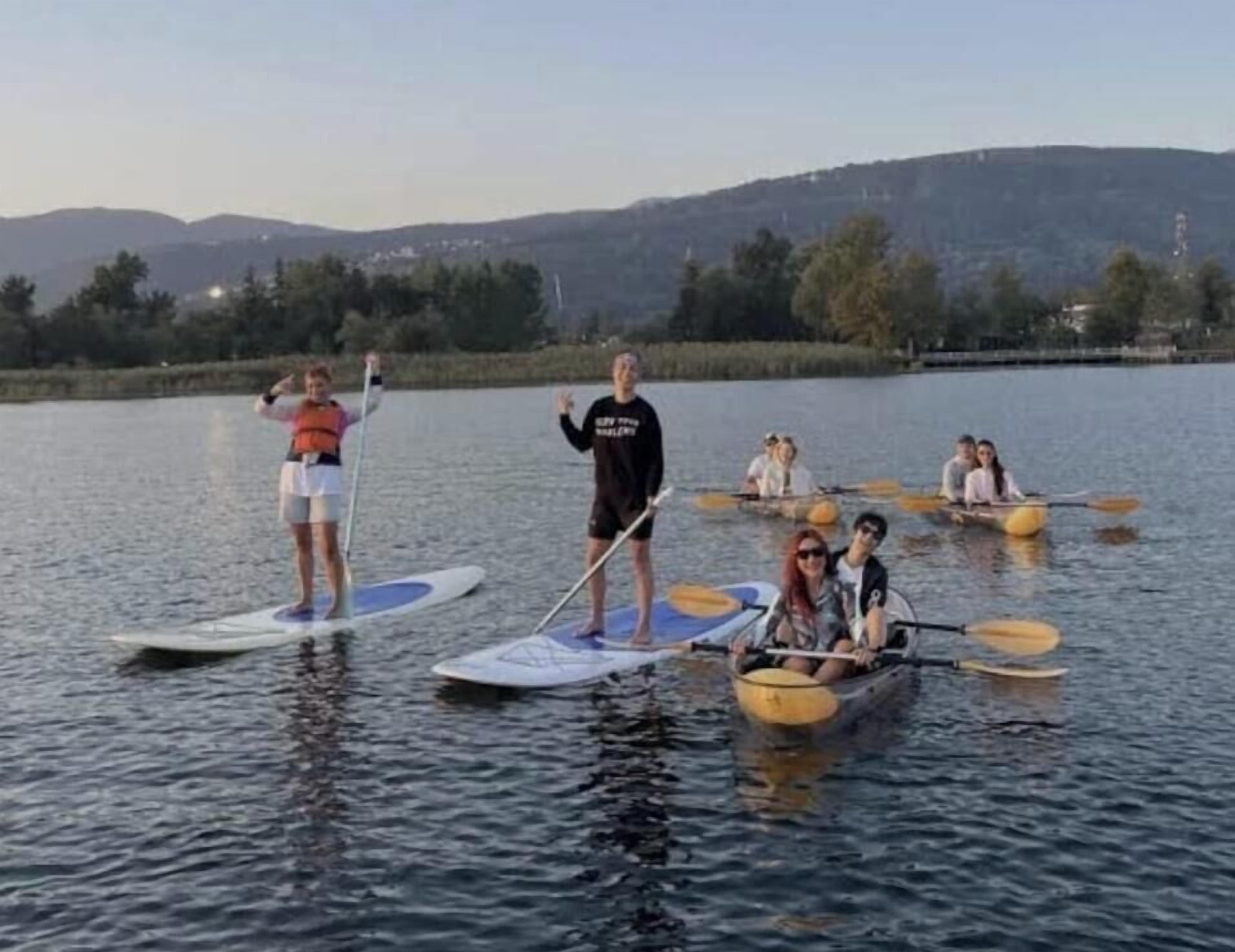 Um grupo de pessoas em pranchas de remo e canoas em um lago calmo, com colinas ao fundo.