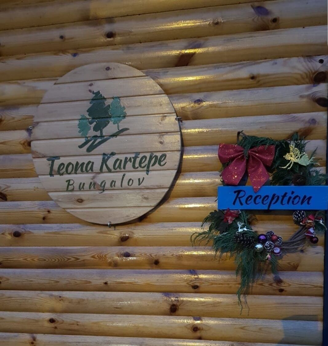 Parede de madeira com uma placa circular que diz 'Teona Kartepe Bungalov' e uma placa azul com a palavra 'Recepção'.
