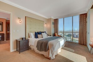 Um quarto espaçoso com uma cama grande, uma mesa de cabeceira, um aparador e vista da cidade através de amplas janelas.