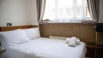 Quarto de hotel com cabeceira de madeira, cama com lençóis brancos, janela com cortinas e mesa de cabeceira com luminária.