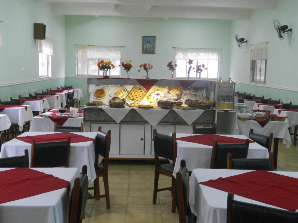 Uma sala de jantar com mesas dispostas em fileiras, cada uma com toalha vermelha. Há arranjos de flores nos parapeitos das janelas e um quadro na parede.