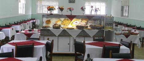 Uma sala de jantar com mesas dispostas em fileiras, cada uma com toalha vermelha. Há arranjos de flores nos parapeitos das janelas e um quadro na parede.