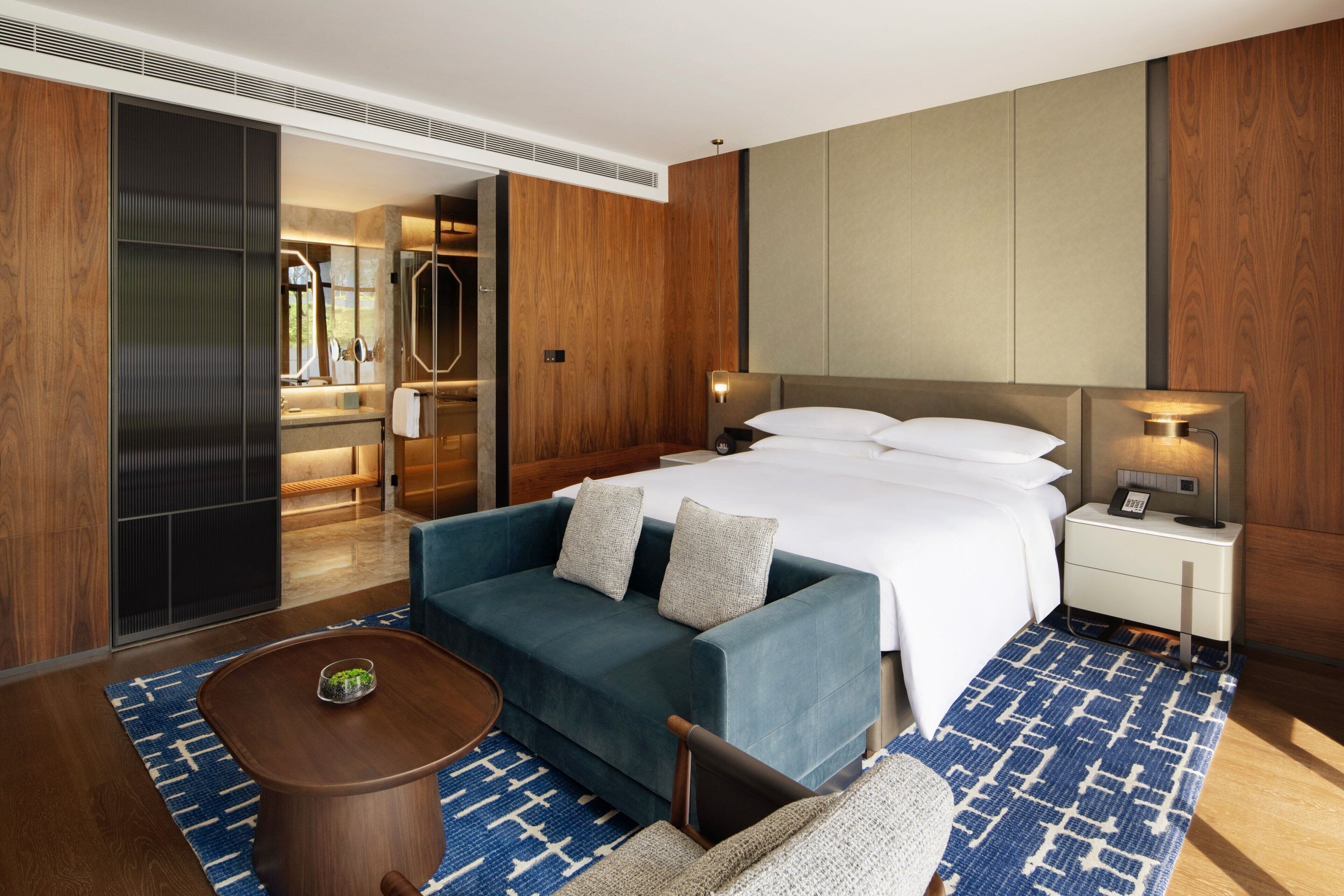 Quarto de hotel com cama, sofá azul, mesa de cabeceira e pequena mesa de centro.