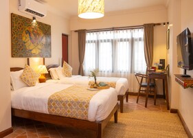 Quarto de hotel com duas camas, uma mesa, uma televisão e uma janela grande com cortinas.
