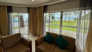 Quarto com sofá e poltronas, janelas amplas com cortinas e vista para uma área gramada e água.
