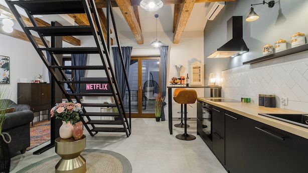 Cozinha moderna com escada de madeira, área de jantar com um banqueta e sala de estar com sofá e plantas decorativas.
