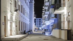 Vista noturna de uma rua estreita de paralelepípedos, ladeada por edifícios modernos com varandas e sinalização iluminada.