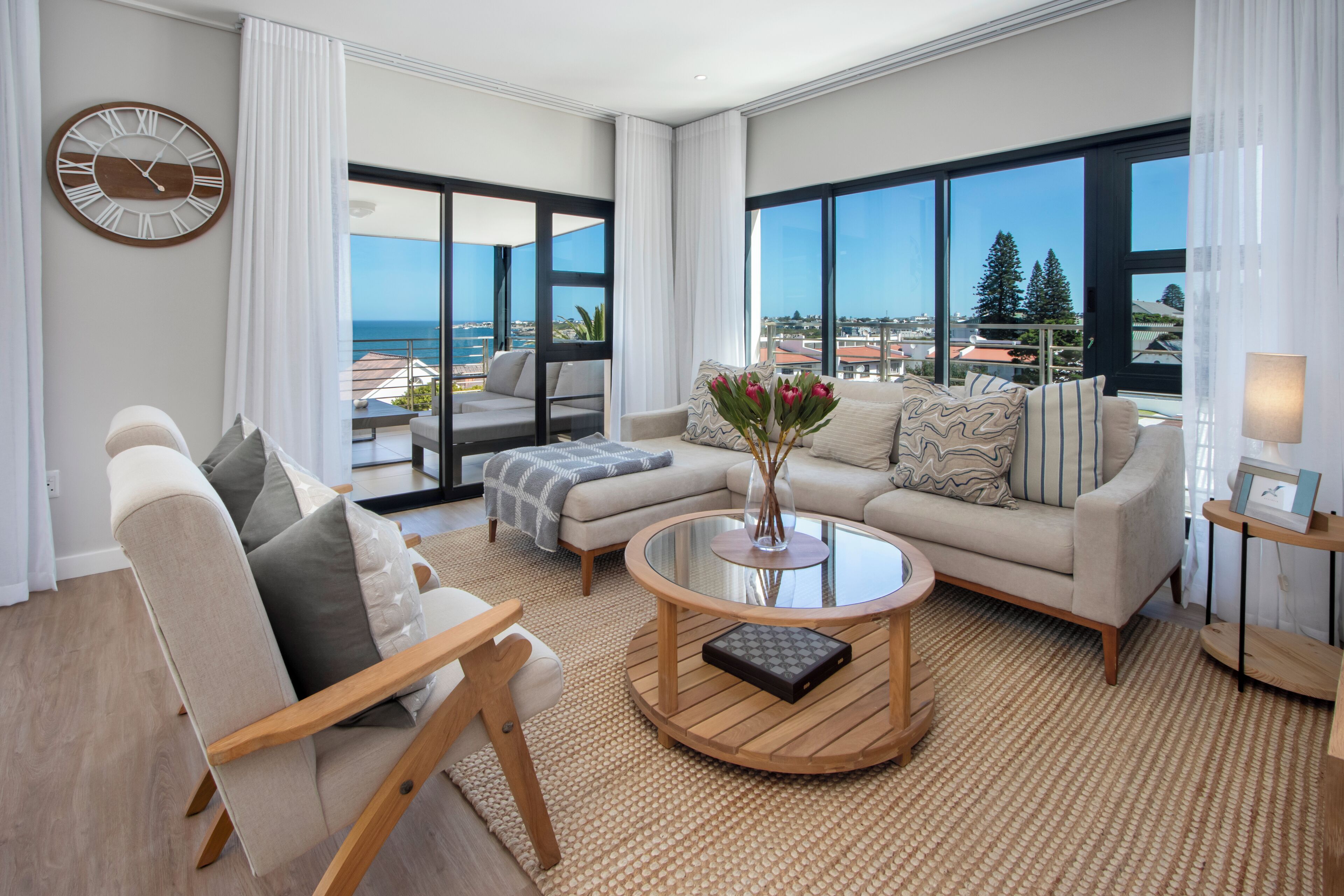 Uma sala de estar moderna com sofá, poltrona e mesa de centro, janelas amplas com vista para o mar e um relógio de parede.