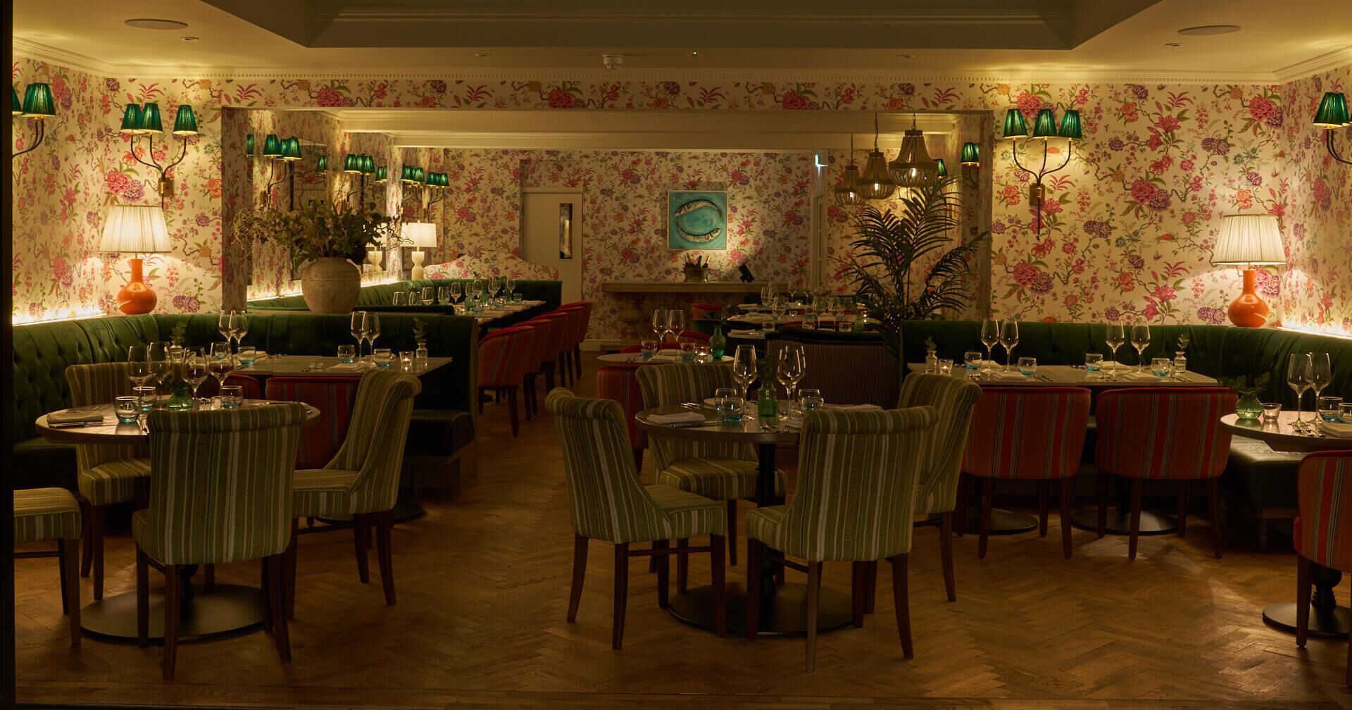 Um restaurante com várias mesas e cadeiras, papel de parede floral e assentos estofados verdes.