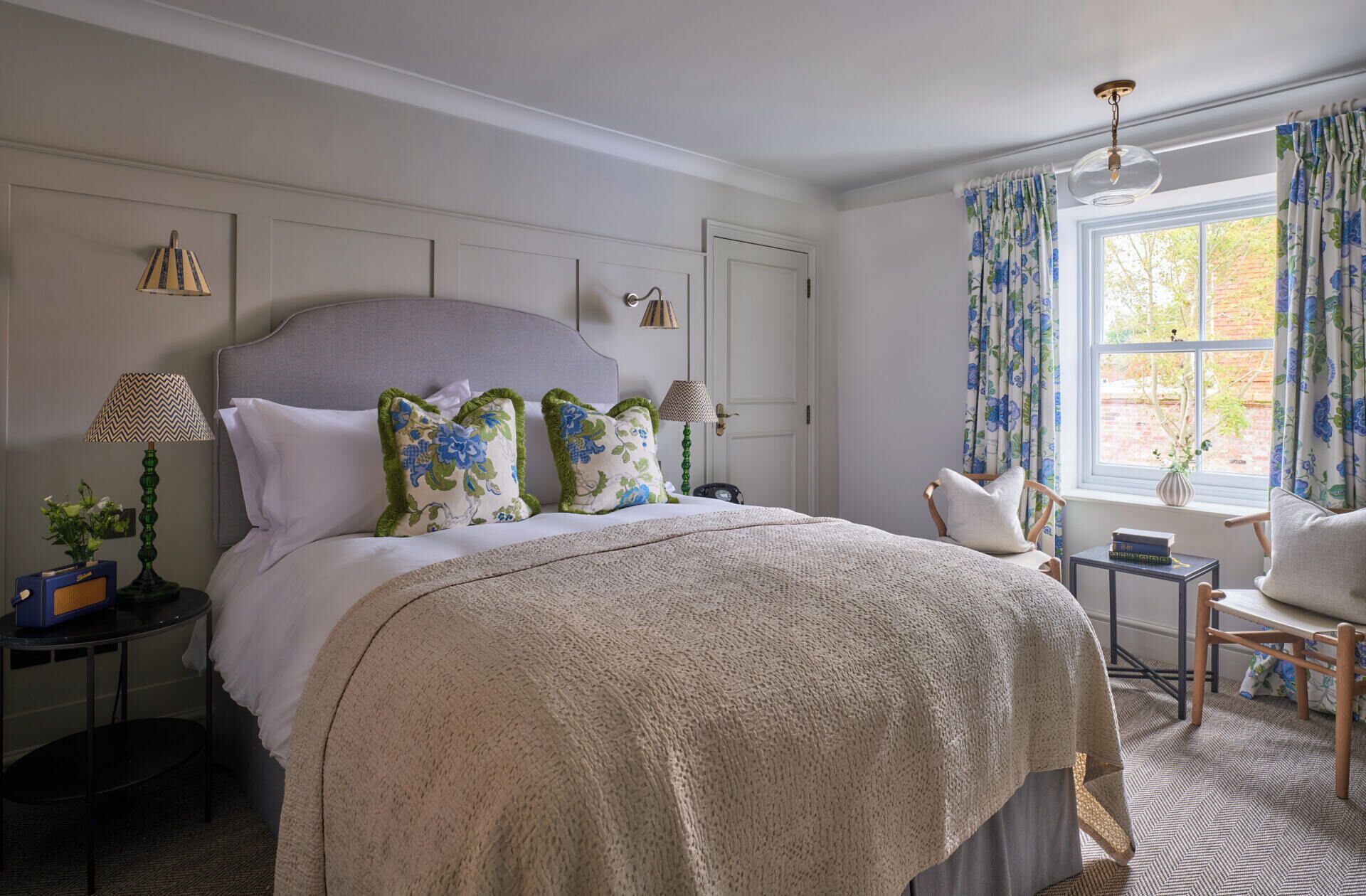 Quarto com uma cama grande, duas luminárias de cabeceira, uma mesa de cabeceira com um rádio, uma cadeira perto da janela e vista para o jardim.