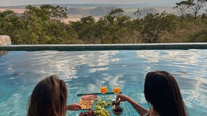 Duas pessoas em mesa à beira da piscina, com variedade de comida e bebidas, apreciando uma paisagem deslumbrante.