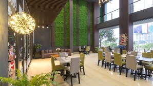 Amplo espaço interno com teto alto, painéis de parede verdes e um grande lustre decorativo. Mesas e cadeiras dispostas para refeições ou reuniões.